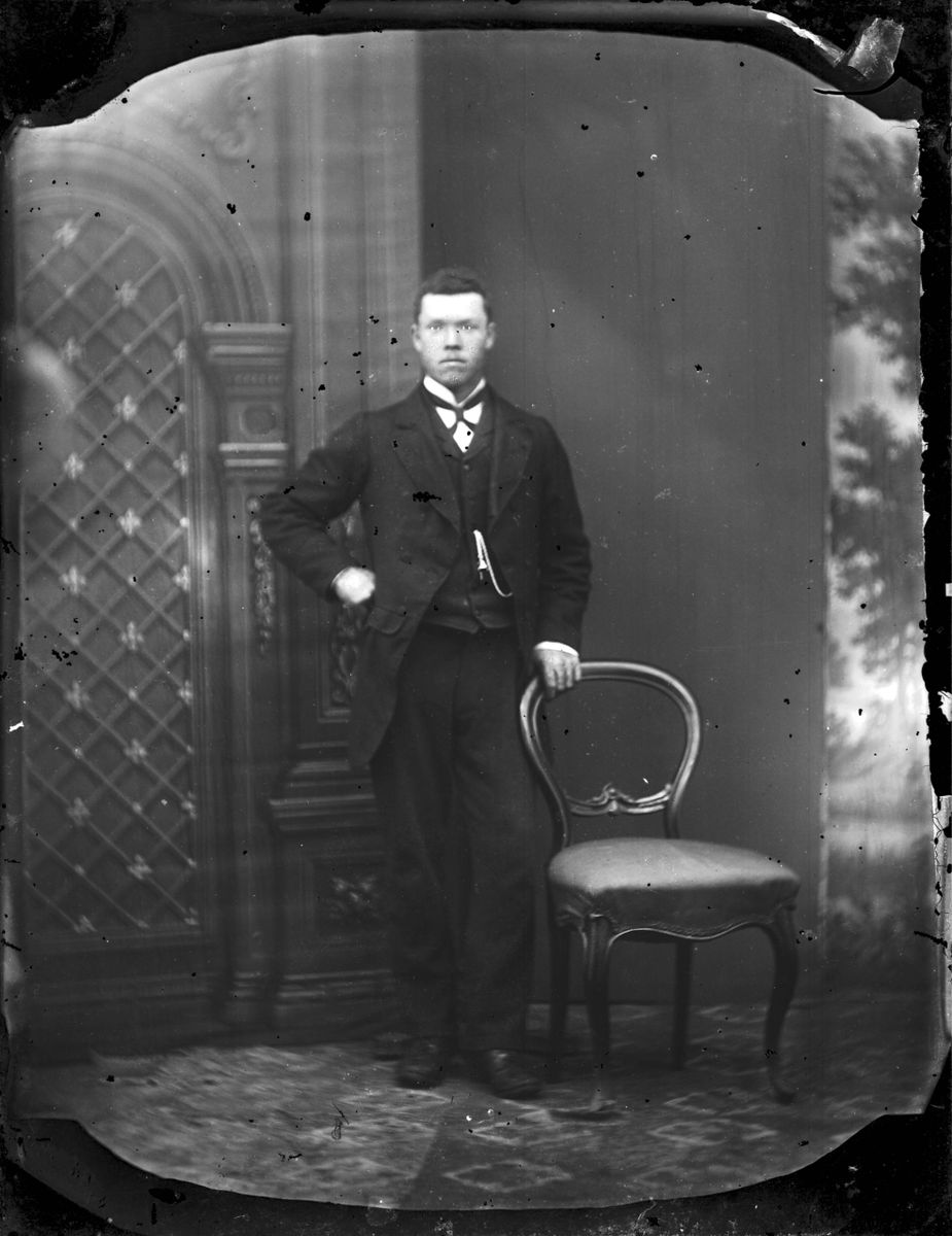 Portrait of a young man, one hand held to the hip, the other on chair ...
